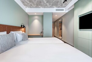 Quarto de hotel moderno com uma cama grande, cabeceira, mesa de cabeceira, televisão e banheiro visível por uma porta aberta.