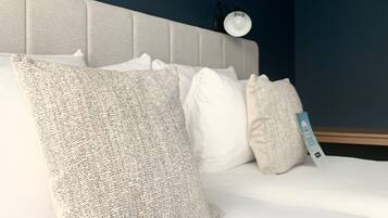Cama bem arrumada com lençóis brancos e várias almofadas, cabeceira estofada cinza e mesa de cabeceira com abajur.
