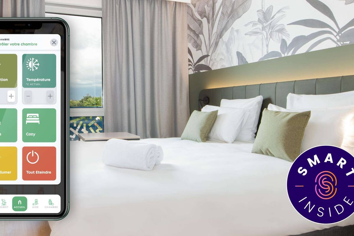 Quarto de hotel com cama, poltrona, televisão e smartphone exibindo um aplicativo de controle do quarto.