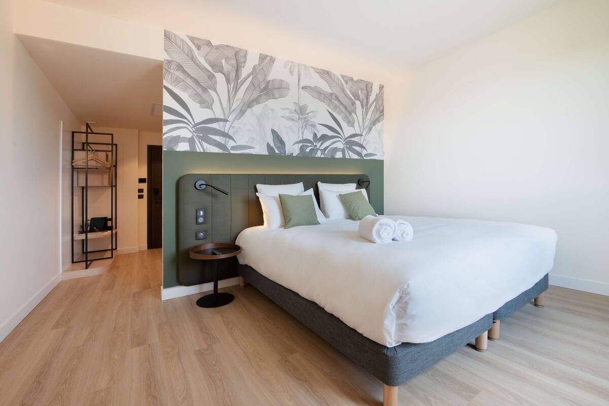 Quarto de hotel moderno com uma cama grande, cabeceira com arte na parede estampada com folhas, mesa de cabeceira e luminária pendente.