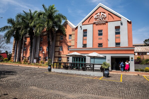 Um hotel com fachada vermelha e cinza, palmeiras e área externa com mesas.