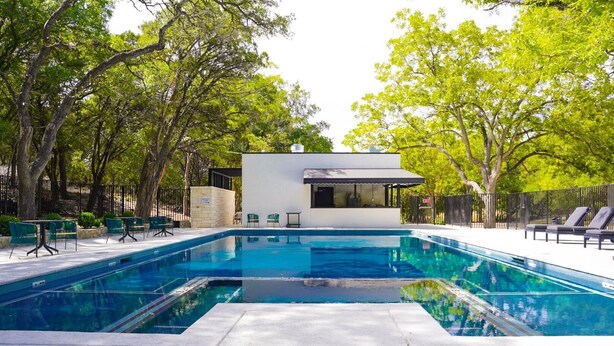 Piscina retangular com um edifício moderno ao fundo, rodeada por árvores e mobiliário para áreas externas.
