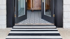 Uma porta com moldura preta e um painel de vidro, um tapete listrado em preto e branco e um armário de madeira com um vaso de flores amarelas.