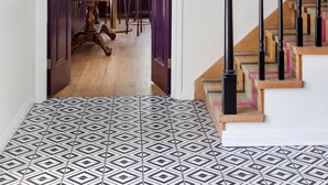 Um corredor com piso estampado, escada com degraus de madeira e corrimãos pretos, e uma área de jantar com mesa e cadeiras.