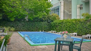 Piscina com um terraço de pedra, cercada por vegetação e um edifício com varandas.