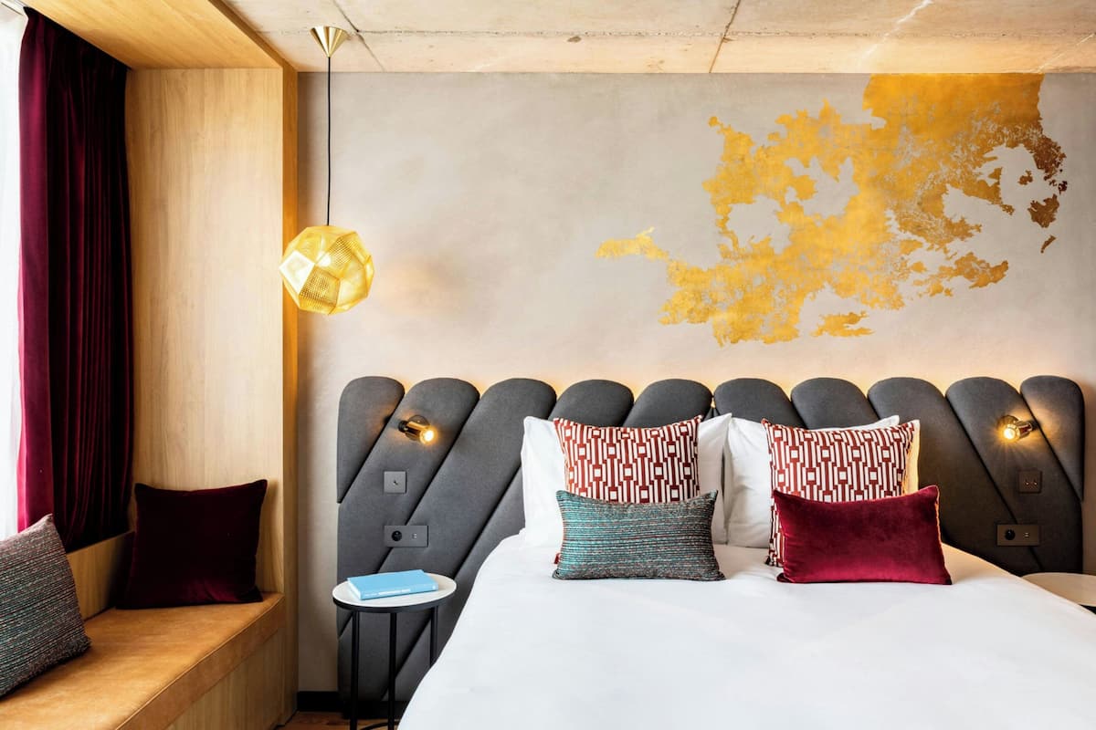 Quarto de hotel com uma cama grande, várias almofadas, uma mesinha de apoio e cabeceira com iluminação embutida.