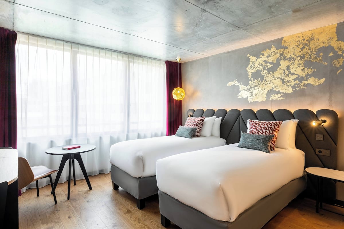 Quarto de hotel com duas camas, uma mesa pequena, uma cadeira e uma janela grande com cortinas.