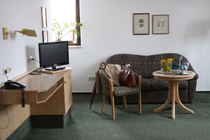 Um quarto com uma mesa, televisão, sofá, mesa redonda pequena e uma planta.