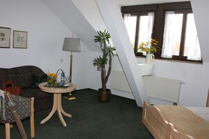 Um quarto pequeno e acolhedor, com uma cama, um sofá, uma mesinha com uma luminária, uma planta em vaso e uma janela com cortinas.