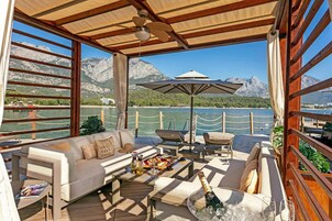 Uma área de estar coberta ao ar livre com sofá, cadeiras, uma mesa com comida e bebida, e vista para um lago e montanhas.