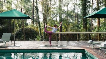 Uma pessoa fazendo uma postura de yoga em um deck de madeira perto de uma piscina, com uma floresta ao fundo.