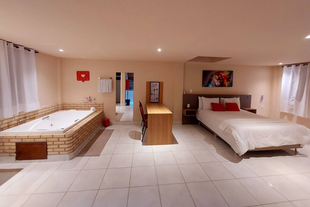 Quarto de hotel moderno com uma banheira grande, cama com almofadas vermelhas e um quadro na parede.