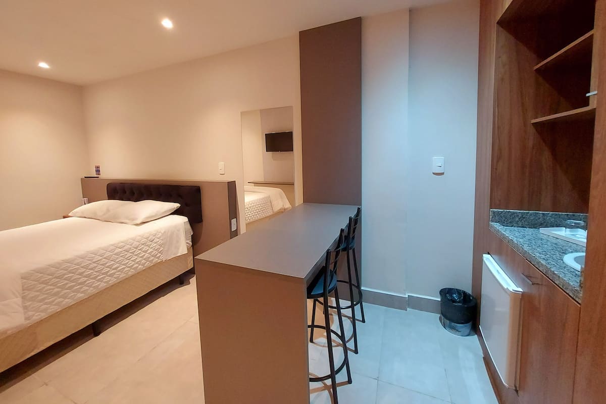Quarto de hotel moderno com uma cama grande, uma mesa com banquetas altas, televisão na parede e uma pequena cozinha.