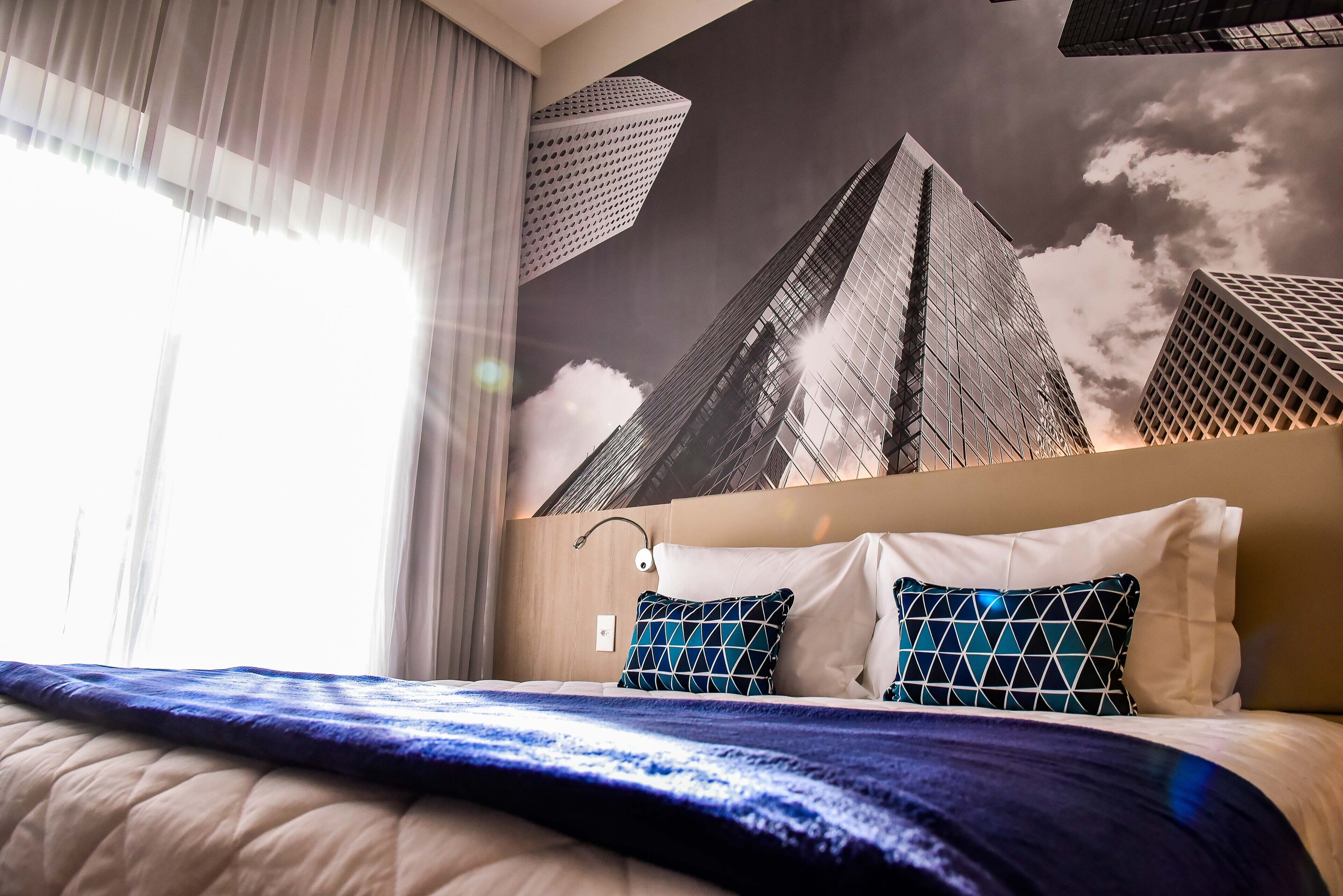 Quarto de hotel com cama, uma janela grande com cortinas leves e um mural na parede com arranha-céus.