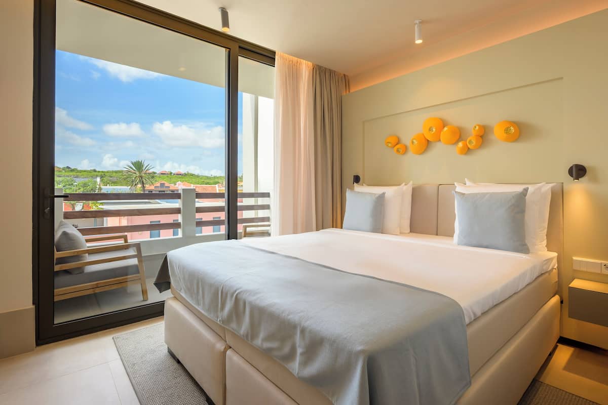 Quarto de hotel com uma cama grande, uma varanda com vista para edifícios e vegetação, e uma parede com decorações circulares.