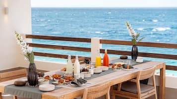 Mesa de jantar de madeira posta com pratos, copos e garrafas, com vista para um corpo d'água.