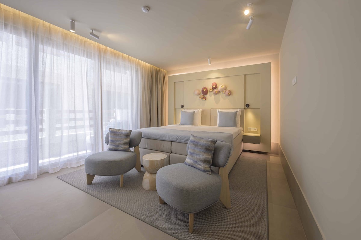 Quarto de hotel moderno com uma cama grande, duas poltronas, uma mesa redonda pequena e um objeto decorativo na parede.