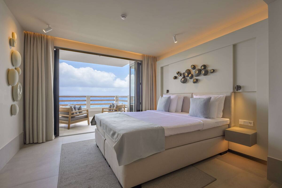 Quarto de hotel moderno com uma cama grande, vista para o mar e varanda com uma cadeira de madeira.