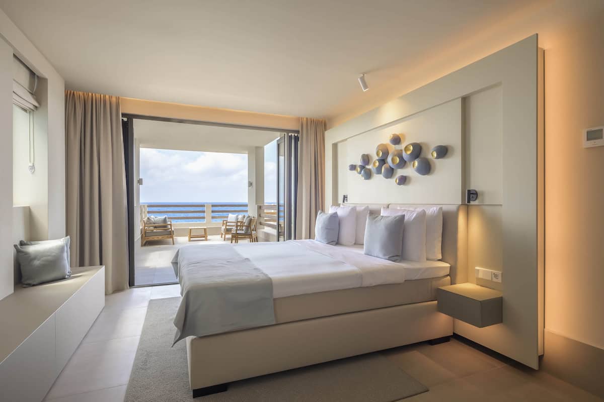 Quarto de hotel moderno com uma cama grande, área de estar e vista para o mar, além de luminária fixada na parede.