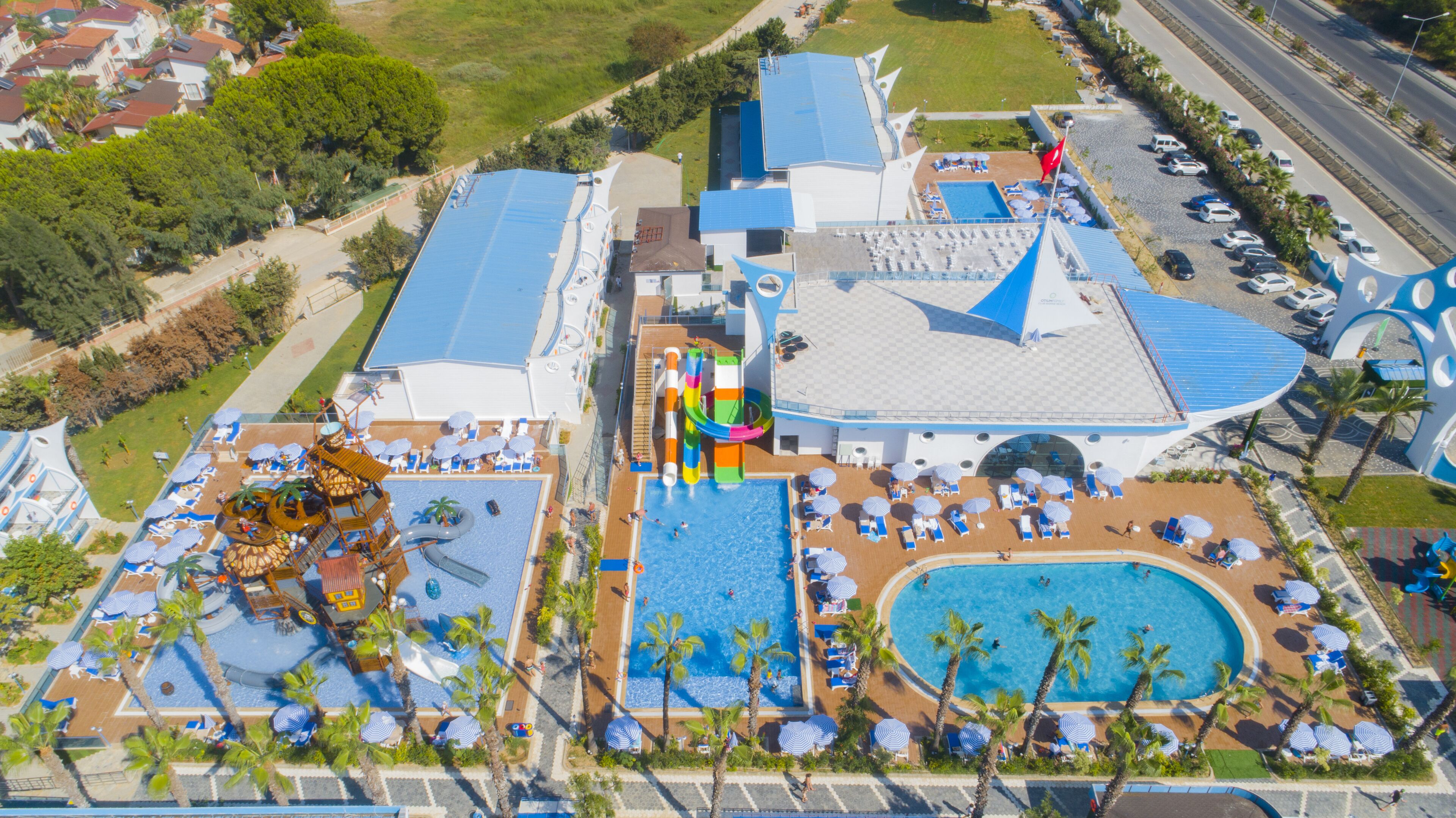 Vista aérea de um resort com uma piscina grande, toboáguas e uma área de praia com espreguiçadeiras e guarda-sóis.