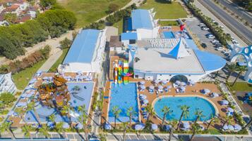 Vista aérea de um resort com uma piscina grande, toboáguas e uma área de praia com espreguiçadeiras e guarda-sóis.