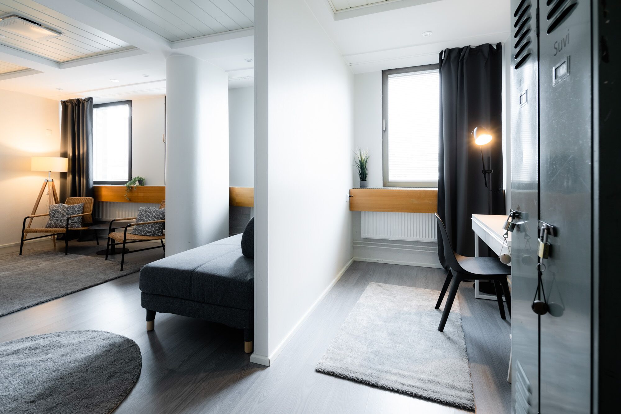 Quarto de hotel moderno com sofá cinzento, uma pequena escrivaninha, uma cadeira e uma grande janela com cortinas.