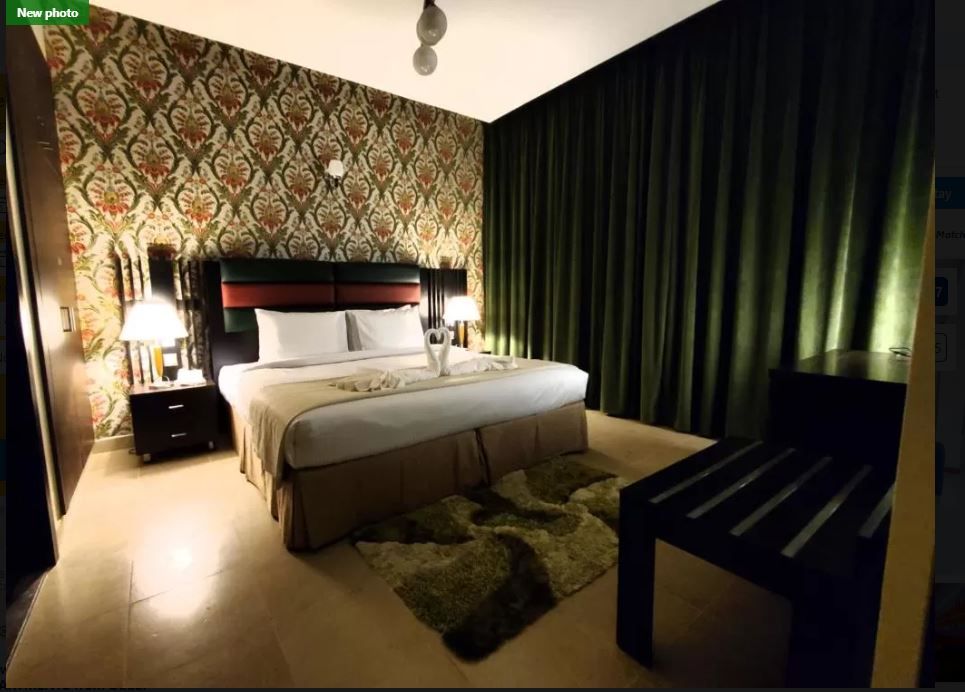 Quarto de hotel com uma cama grande, duas mesas de cabeceira com luminárias, cortina verde e papel de parede estampado.