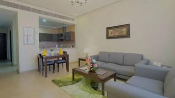 Uma sala de estar com sofá, mesa de jantar com cadeiras, mesa de centro com flores e cozinha ao fundo.