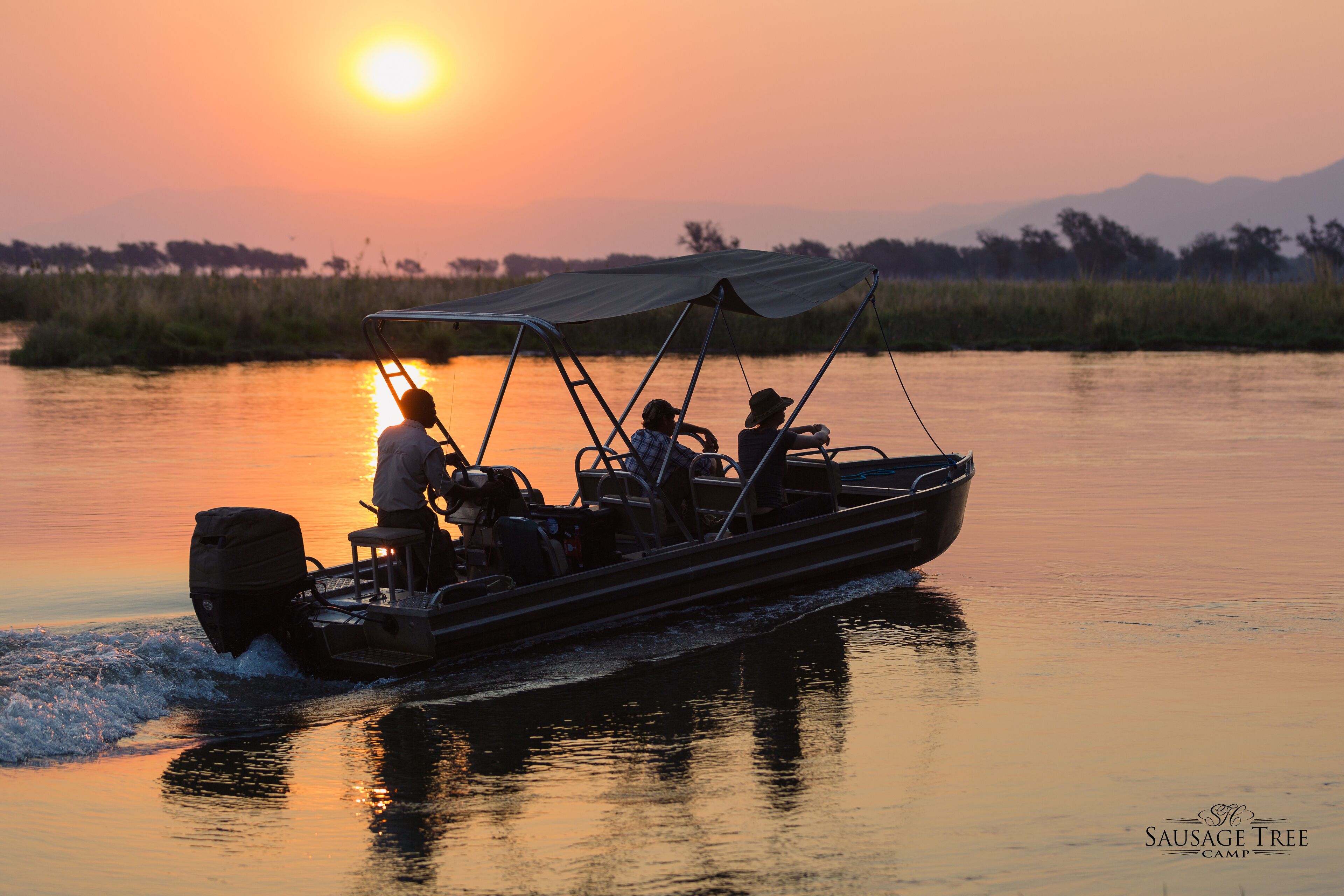 Um barco com toldo, duas pessoas e motor, navegando em águas calmas durante o pôr do sol.
