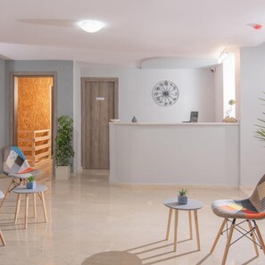 Uma área de recepção com porta de madeira, um relógio de parede e duas cadeiras com pequenas mesas.
