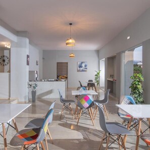 Uma zona de refeições moderna, com mesas e cadeiras, uma cozinha ao fundo e uma janela ampla que oferece luz natural.