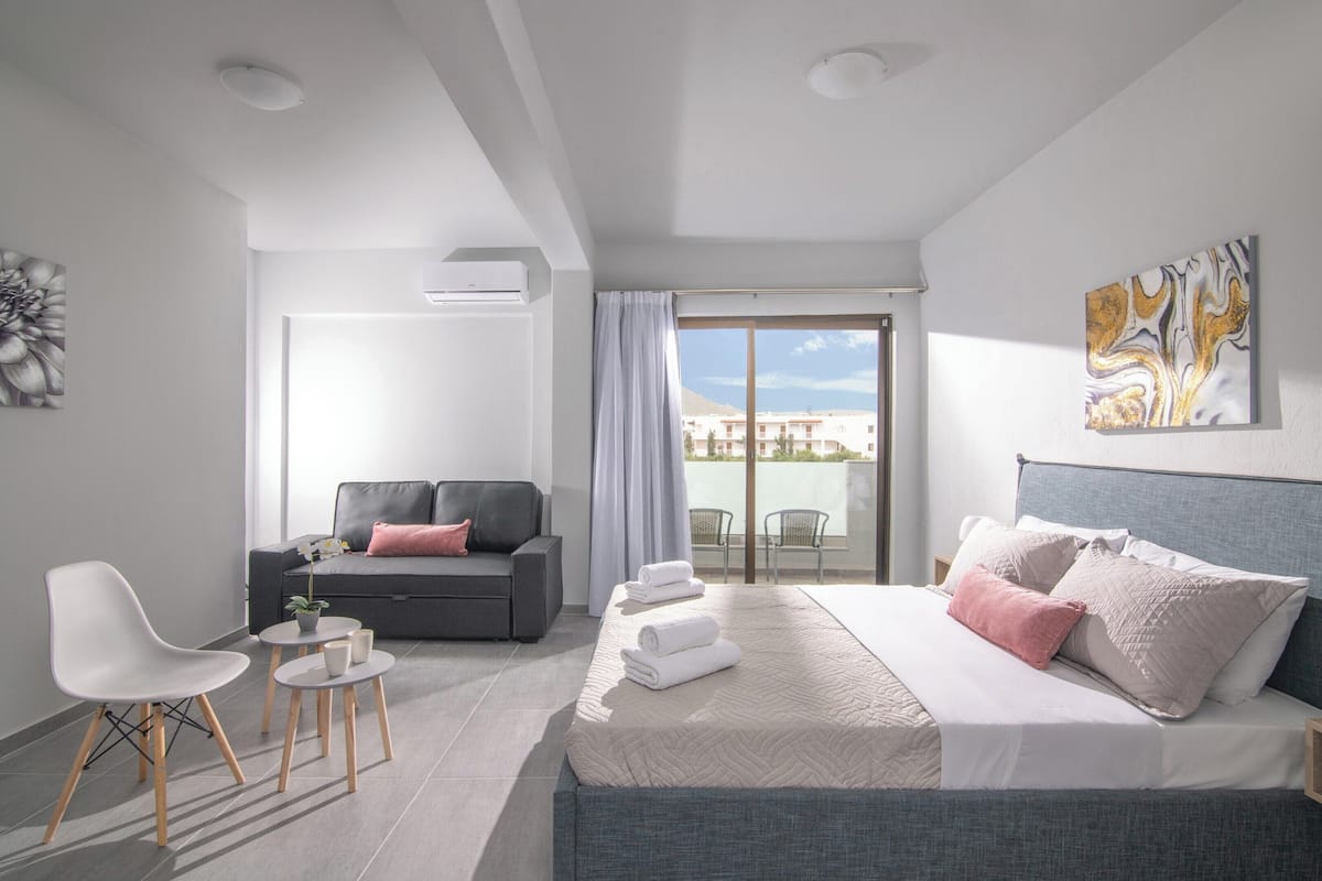 Quarto de hotel moderno com cama, sofá, poltrona, mesa pequena e varanda com vista.