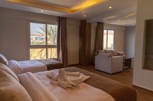 Quarto de hotel com duas camas, um sofá e uma janela grande com vista para palmeiras e edifícios.