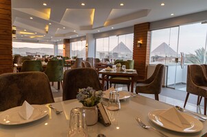 Restaurante com mesas preparadas para refeições, grandes janelas com vista para uma pirâmide e design de interiores moderno.