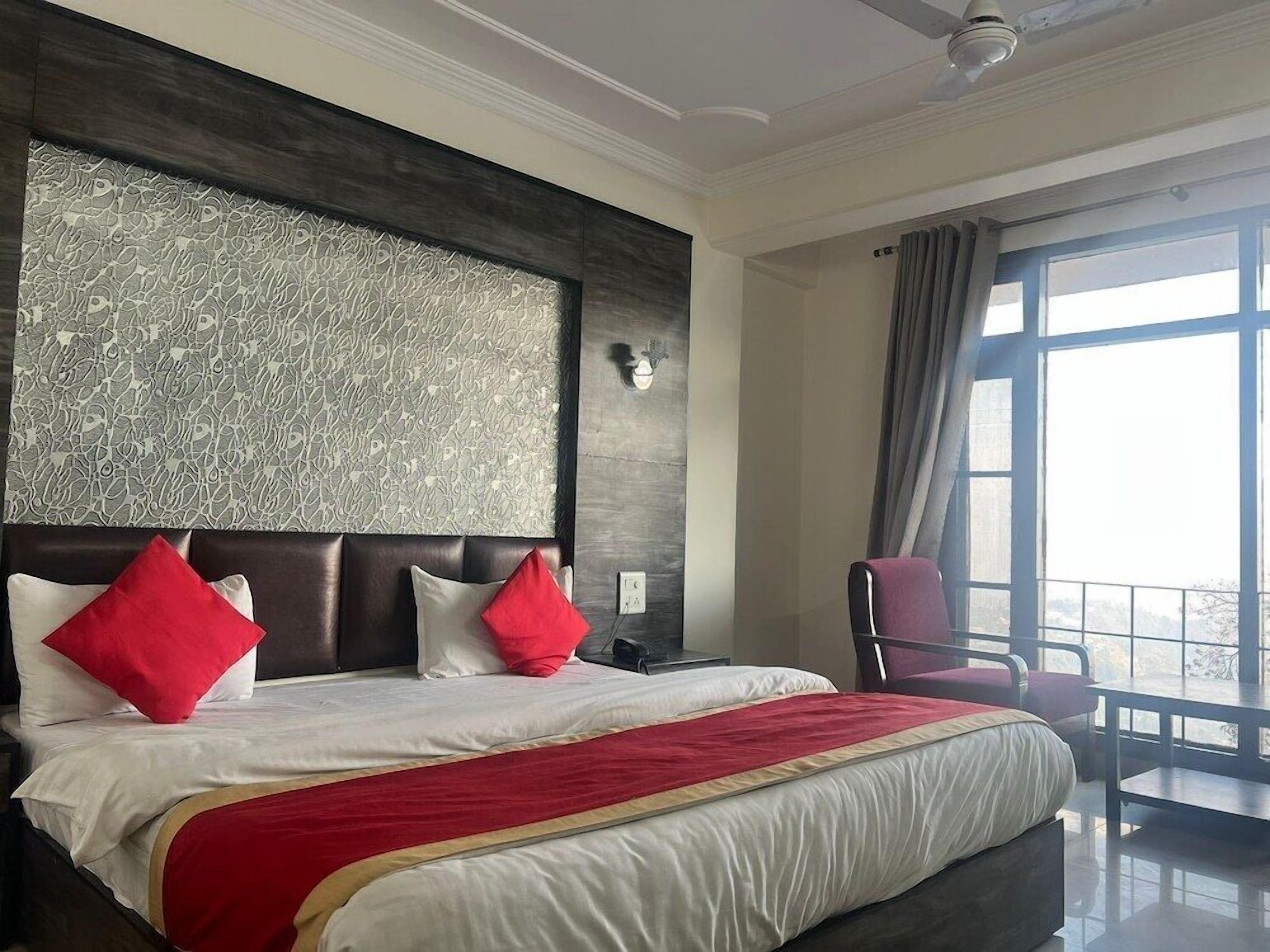 Quarto de hotel com uma cama grande, colcha vermelha e branca, cabeceira escura e uma janela ampla com cortinas.