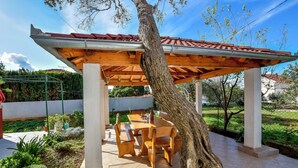 Uma área de refeições ao ar livre coberta, com uma mesa e cadeiras de madeira, cercada por vegetação e um chão de azulejos.