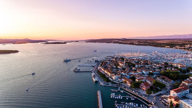 Uma cidade costeira com um porto repleto de barcos, rodeada por edifícios residenciais e uma paisagem montanhosa ao fundo.