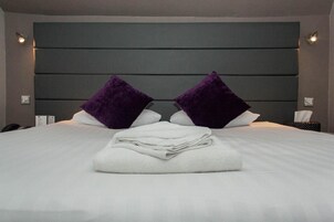 Cama bem arrumada com lençóis brancos e duas almofadas roxas, uma cabeceira escura e luminárias de cabeceira.
