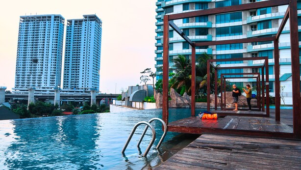 Piscina com deck de madeira e escada de metal, rodeada por um edifício moderno com fachada azul e branca.