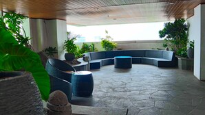 Uma área de estar moderna ao ar livre, com um sofá curvo e uma mesa redonda, rodeada por plantas em vasos e um chão de lajes de pedra.