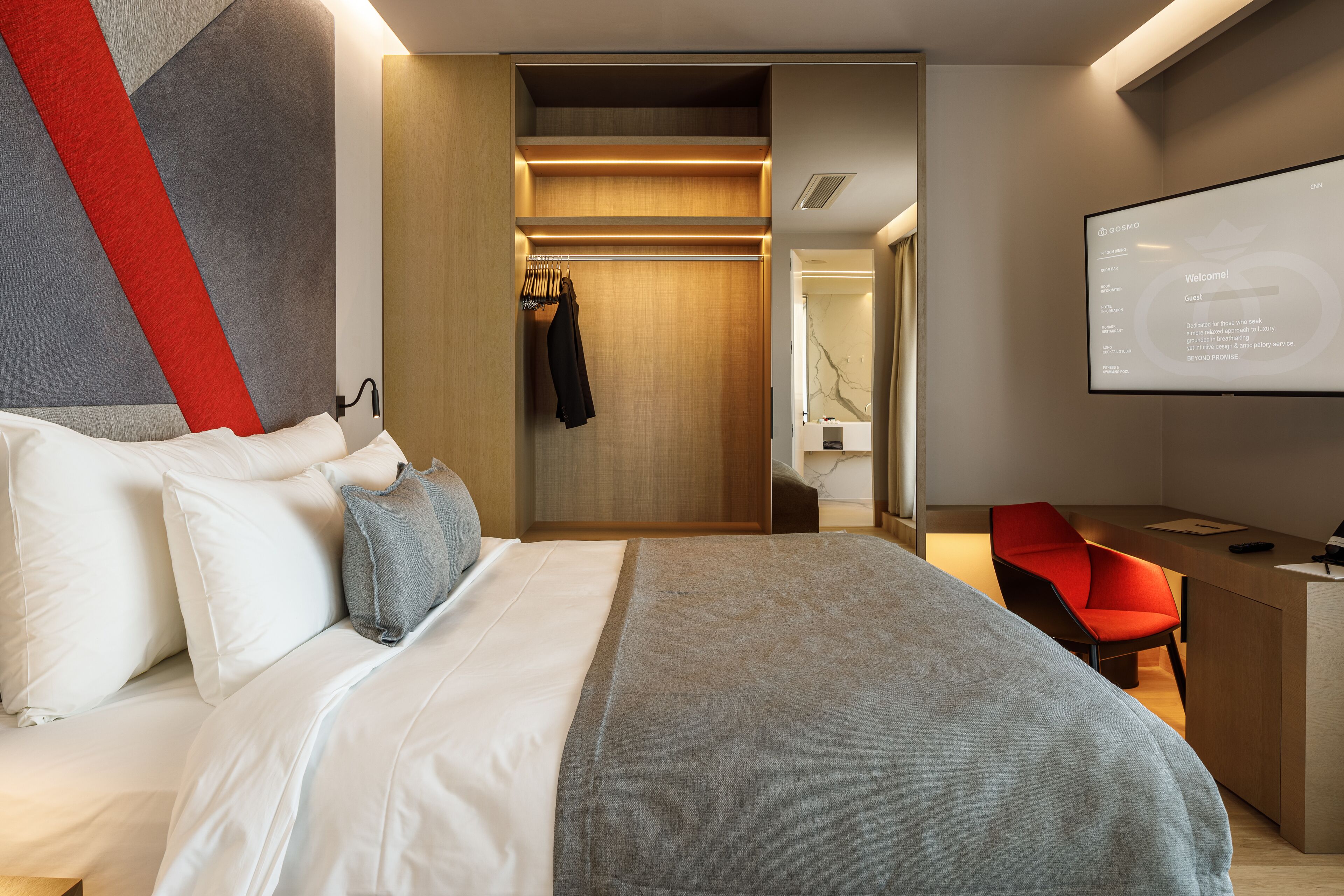 Quarto de hotel moderno com uma cama grande, uma escrivaninha, uma cadeira vermelha e uma TV exibindo uma mensagem de boas-vindas.