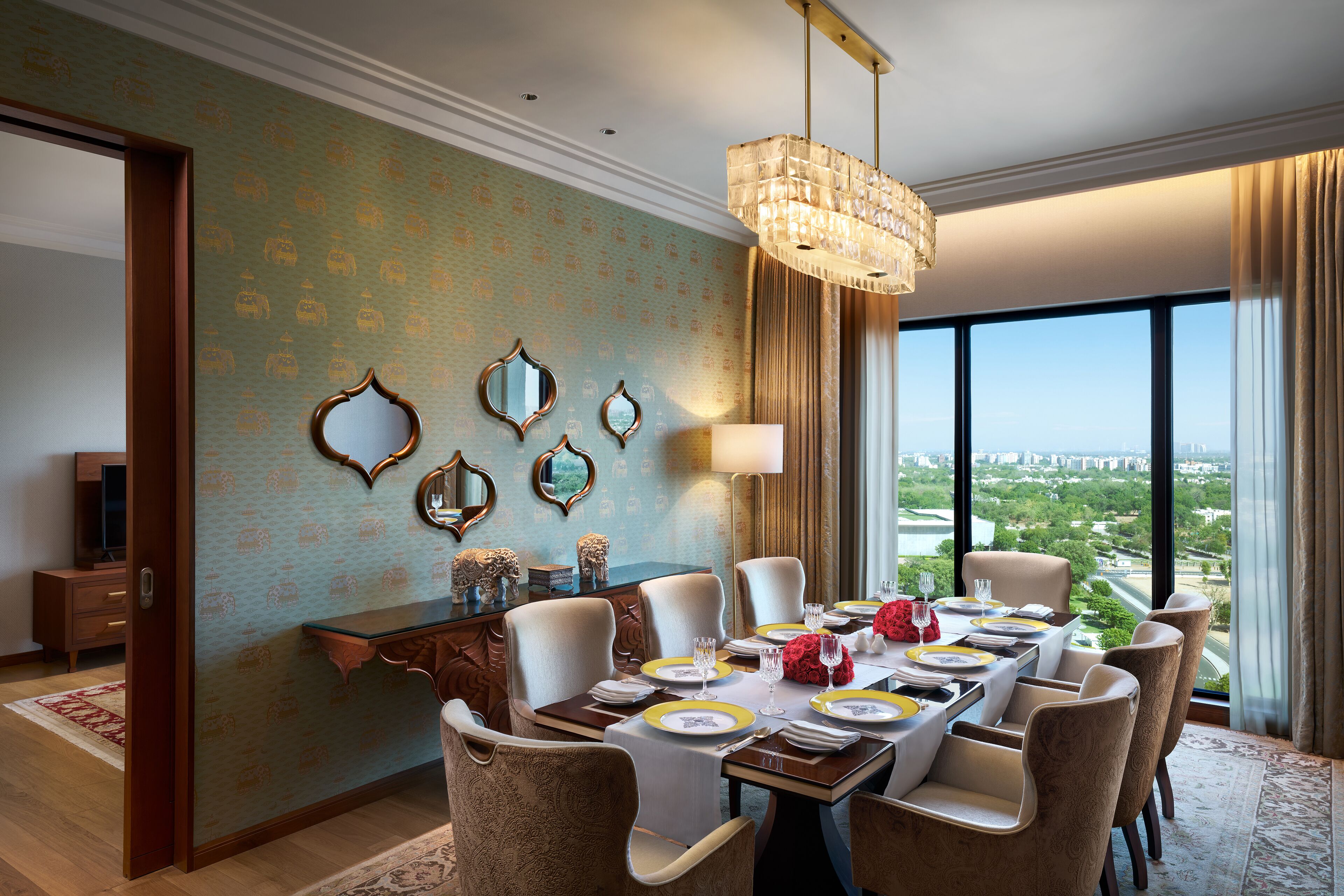 Uma sala de jantar com uma mesa grande posta para seis pessoas, rodeada de cadeiras, com vista para a cidade através de grandes janelas e um lustre acima.