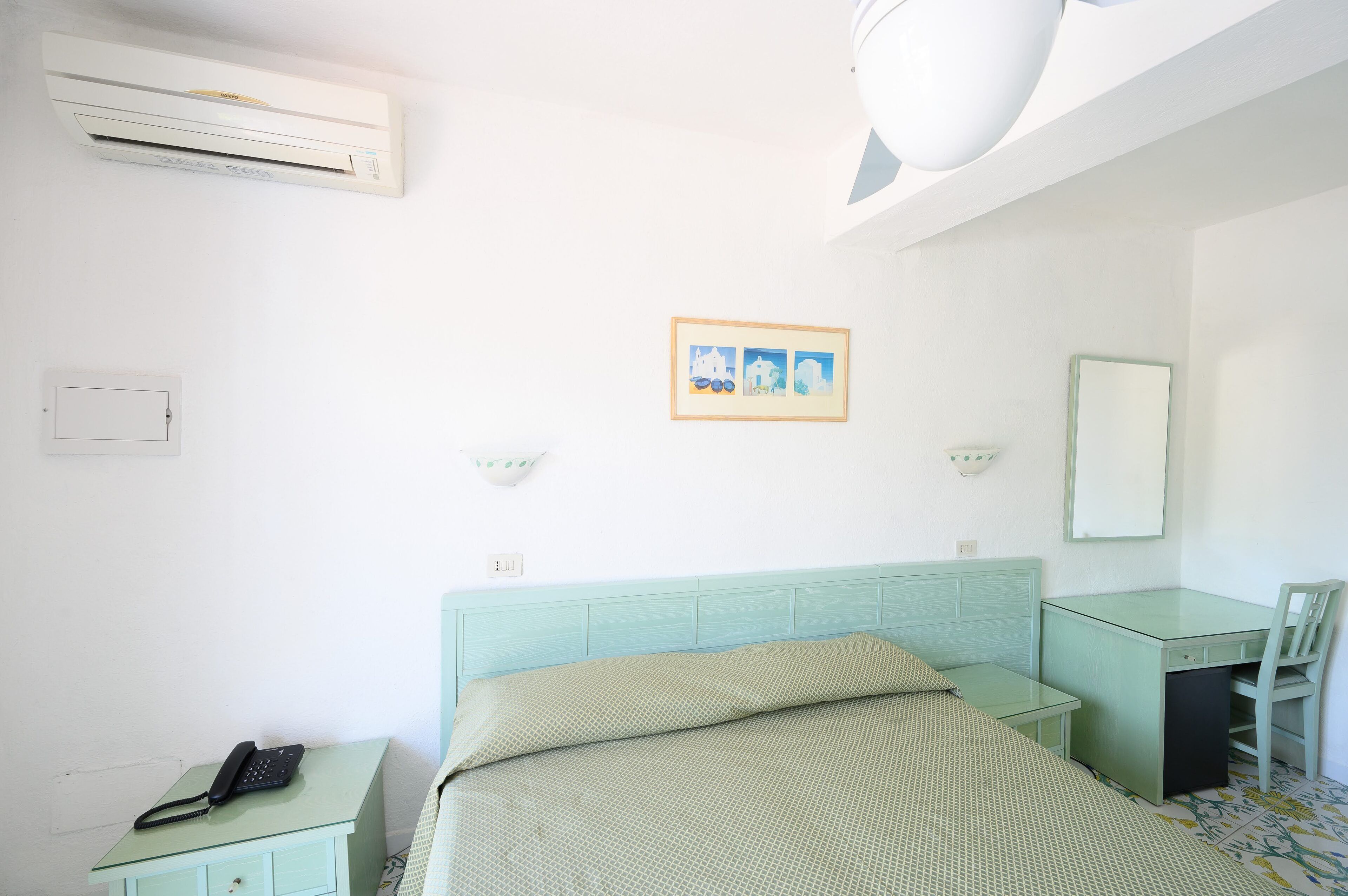 Quarto com cama, escrivaninha, cadeira, ar-condicionado, telefone, quadro emoldurado e espelho.