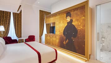Quarto de hotel com uma cama grande, cadeiras estofadas em vermelho, uma mesa redonda pequena e uma grande obra de arte emoldurada na parede.