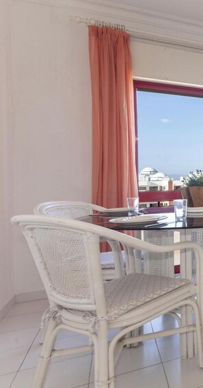 Uma sala de jantar com cadeiras de vime branco e uma mesa de vidro, uma cortina vermelha e vista para edifícios pela janela.
