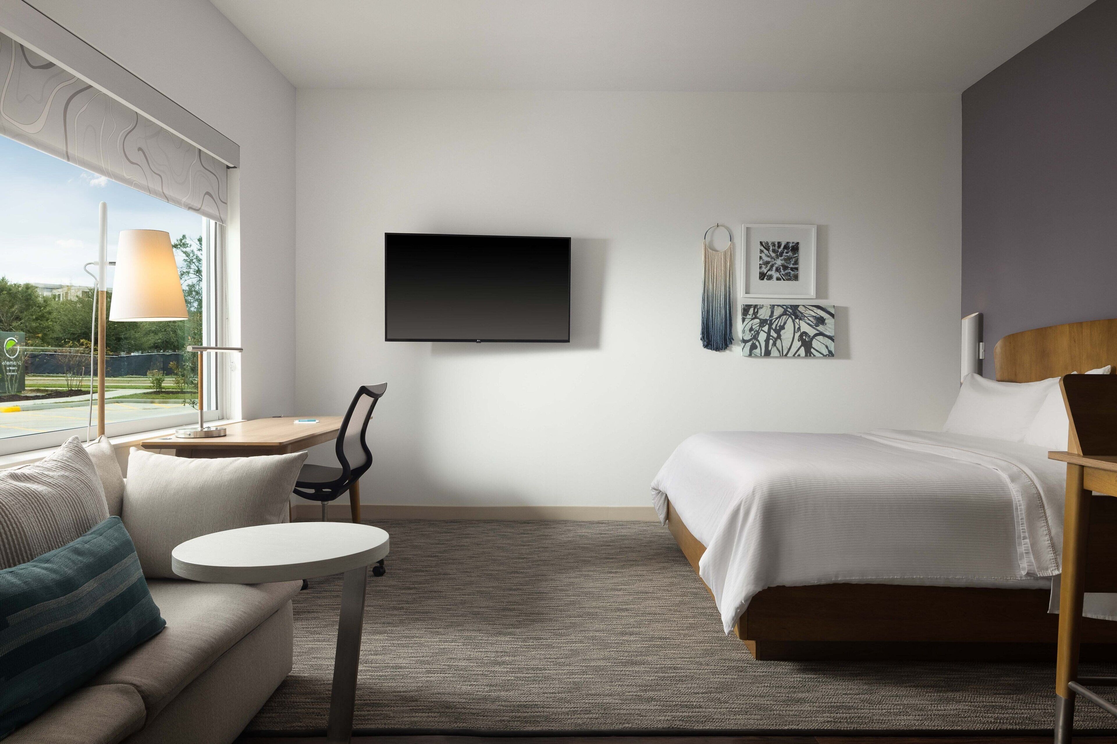 Quarto de hotel moderno com cama, sofá, mesa pequena, televisão e obras de arte na parede.