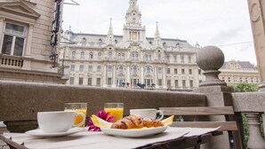Uma mesa com um croissant, uma xícara e um copo de sumo de laranja, com um edifício histórico e arquitetura ornamentada ao fundo.