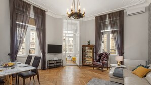 Uma sala de estar espaçosa com piso de madeira em padrão herringbone, um sofá, uma mesa de jantar com cadeiras, uma televisão, uma estante e um lustre.