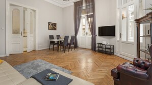 Uma sala de estar ampla com piso de madeira, um sofá, uma poltrona de couro, um espaço de refeições com cadeiras, uma TV e um lustre.