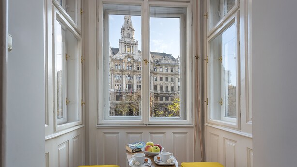 Quarto com vista para edifícios históricos através de amplas janelas, uma pequena mesa com uma tigela de frutas e xícaras, e cadeiras amarelas.